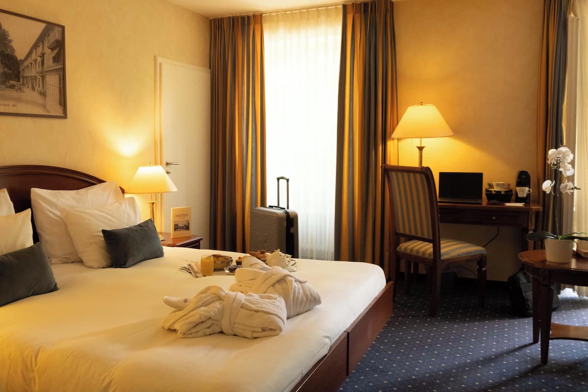 Quarto de hotel com cama, duas luminárias de cabeceira, uma escrivaninha com um laptop, uma cadeira, uma mala e uma mesinha com abajur e flores.