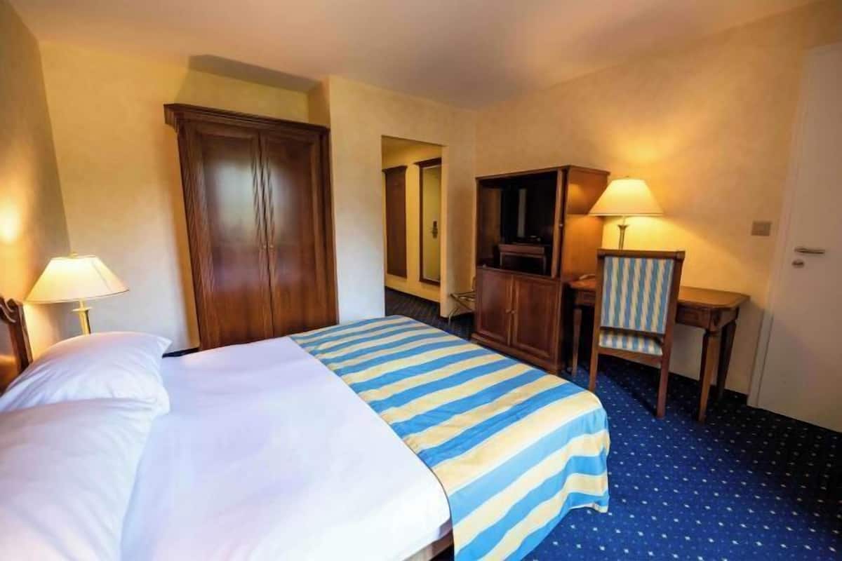 Quarto de hotel com uma cama, escrivaninha, cadeira, televisão e guarda-roupa.