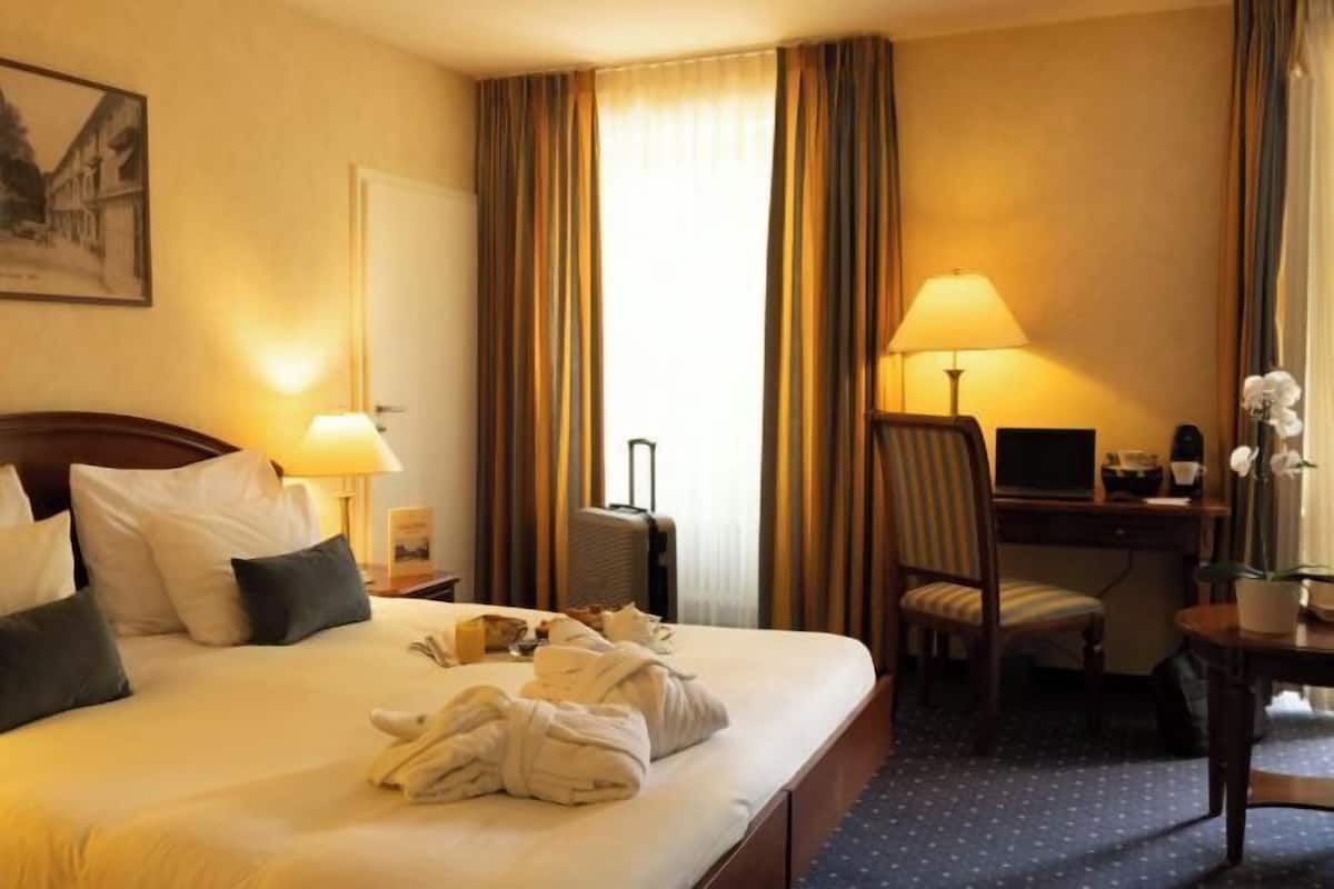 Quarto de hotel com cama, escrivaninha, cadeira, abajur, mala e pequena mesa com planta.