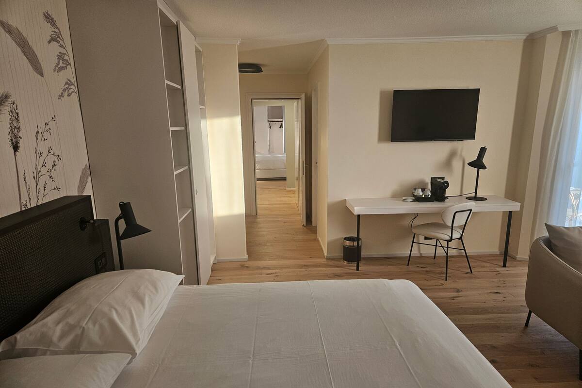 Quarto de hotel com uma cama, uma escrivaninha com cadeira, uma televisão e uma janela.