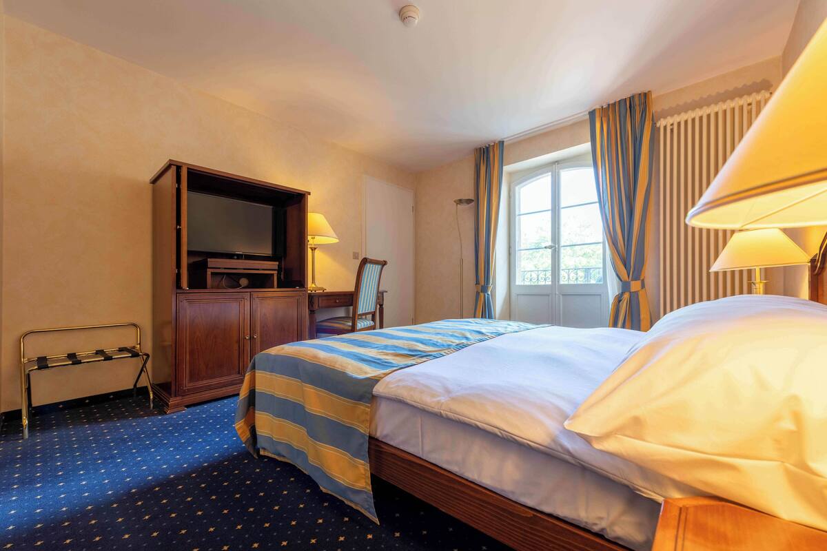 Quarto de hotel com cama, televisão, escrivaninha e janela com cortinas.