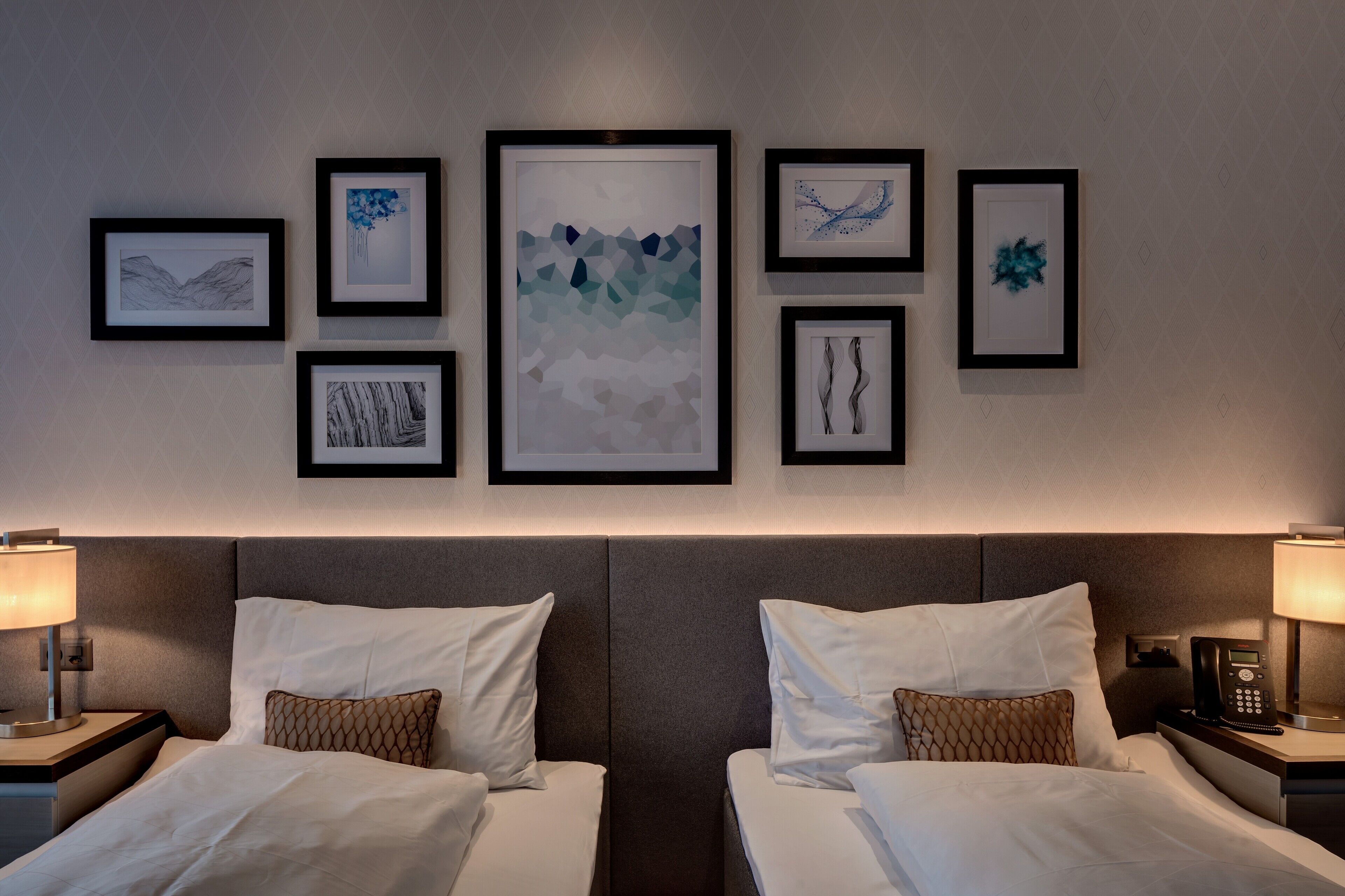 Quarto de hotel com duas camas, cabeceiras, mesas de cabeceira e quadros nas paredes.