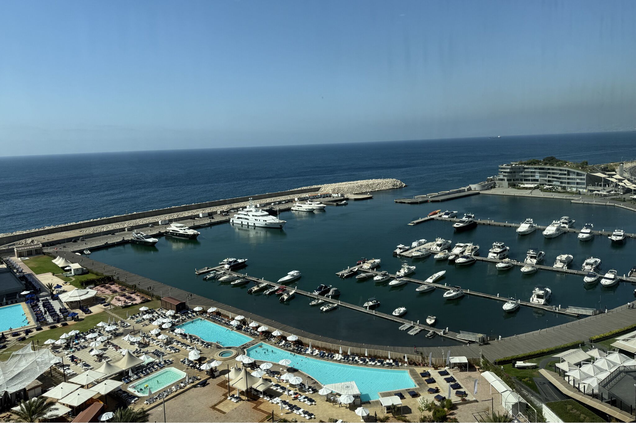 Uma marina com vários barcos atracados, uma área de piscina com guarda-sóis e um edifício com telhado curvo.