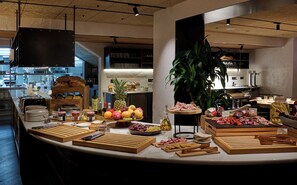 Buffet montado com diversos alimentos, incluindo carnes, queijos e frutas, numa cozinha bem iluminada com eletrodomésticos modernos e uma grande planta.