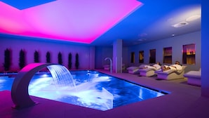 Uma área interna de piscina moderna, com cachoeira, espaços para sentar e teto com iluminação geométrica.