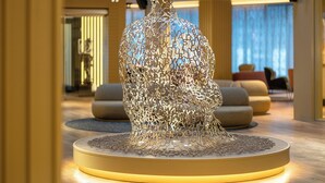 Escultura de uma figura humana feita de letras, colocada em uma plataforma circular em um hall moderno com áreas de estar e uma grande janela.