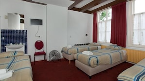 Quarto de hotel com duas camas, uma cadeira vermelha, uma mesinha com uma pia branca e uma janela com cortinas vermelhas.