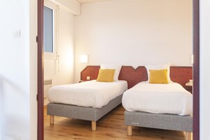 Quarto com duas camas, cada uma com estrutura cinza e roupa de cama branca, e chão de madeira.