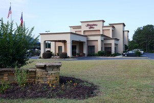 Um hotel Hampton Inn com fachada bege, bandeiras americanas e um jardim frontal ajardinado com flores e arbustos.