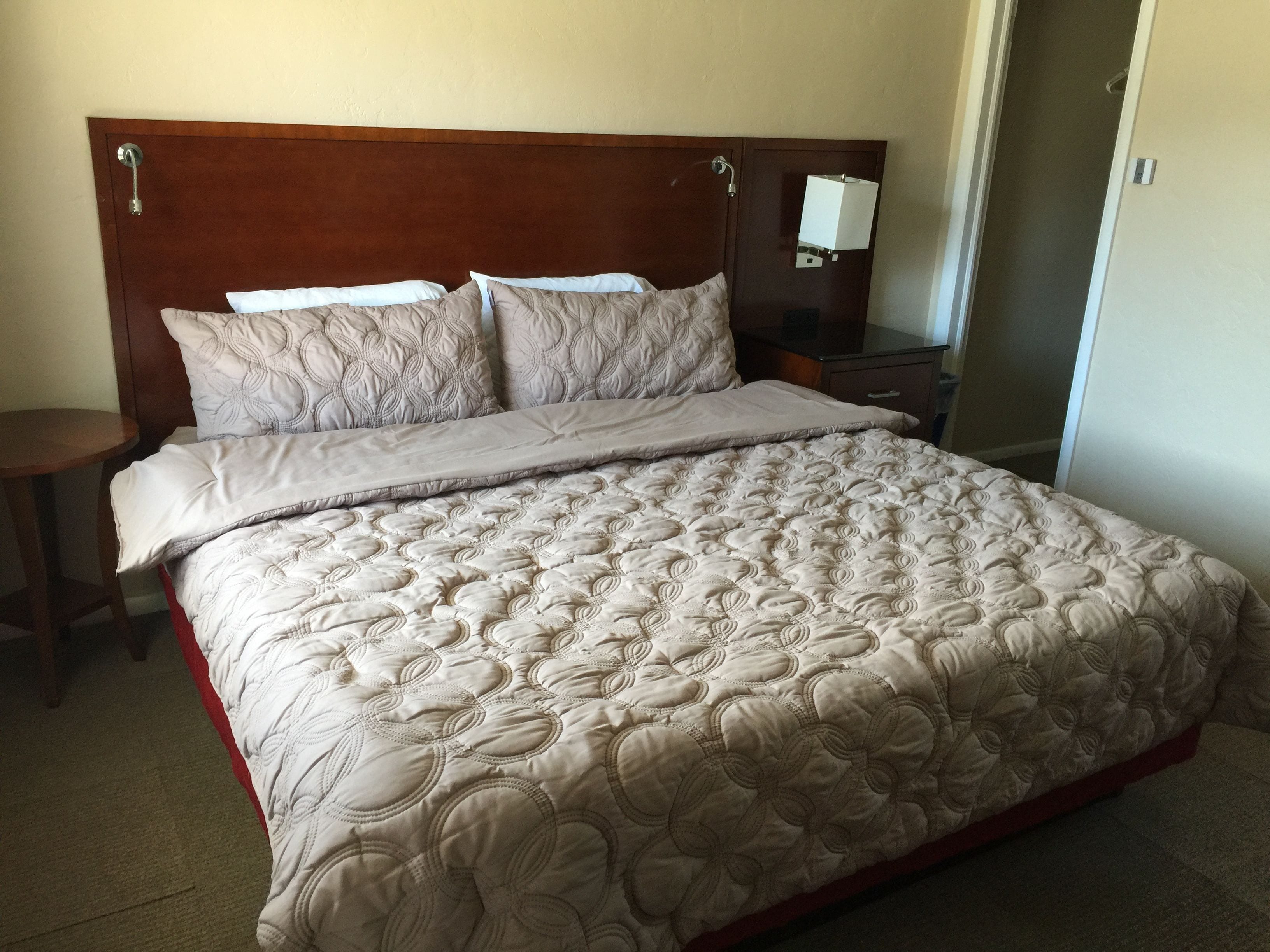 Cama bem arrumada com colcha estampada e almofadas, cabeceira de madeira, mesa de cabeceira com abajur e criado-mudo.