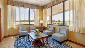 Quarto de hotel com uma janela grande, mesa de centro de madeira, duas poltronas estampadas, mesinha lateral com luminária e tapete azul estampado.
