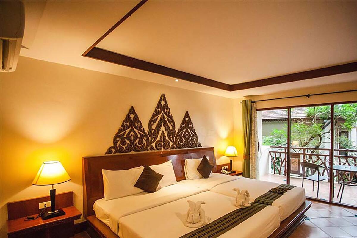 Quarto de hotel com uma cama grande, duas luminárias de cabeceira, um painel decorativo e uma varanda com mobiliário de exterior.