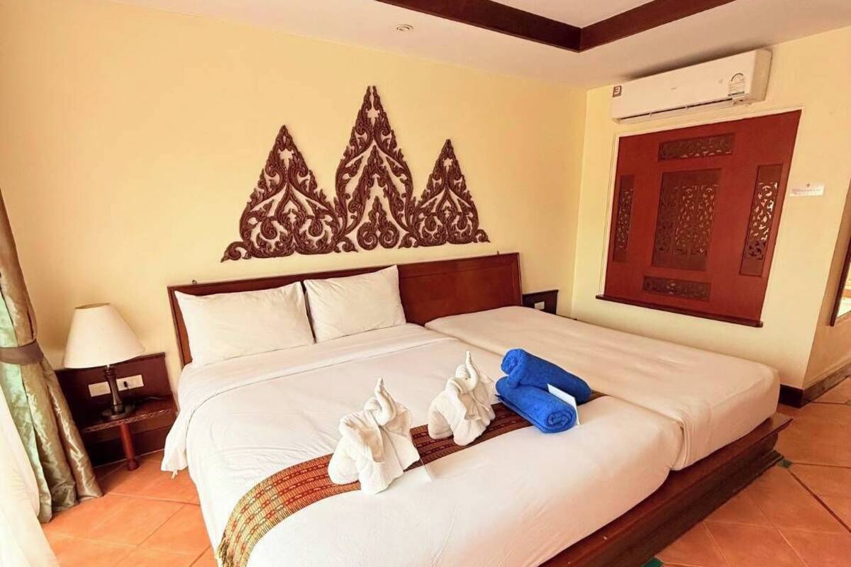 Quarto de hotel com uma cama grande, cabeceira decorativa, mesa de cabeceira com abajur e uma porta vermelha.