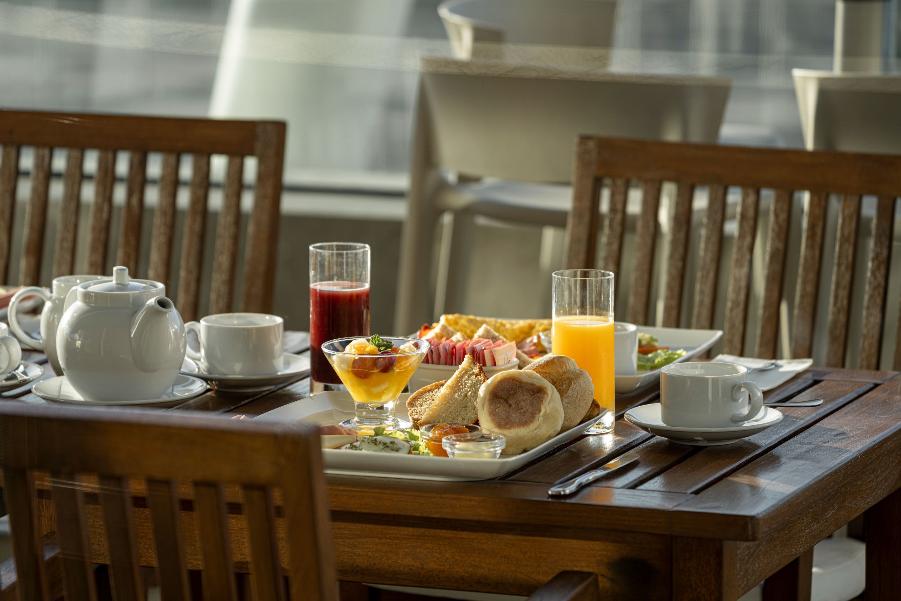Uma mesa de madeira posta para o café da manhã, com cadeiras, um bule, xícaras, um prato com ovos, torradas e suco de laranja.