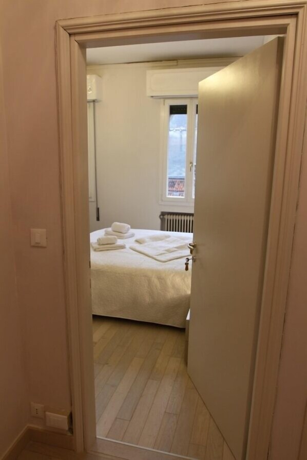 Corredor estreito que dá para um quarto com cama, radiador e janela.