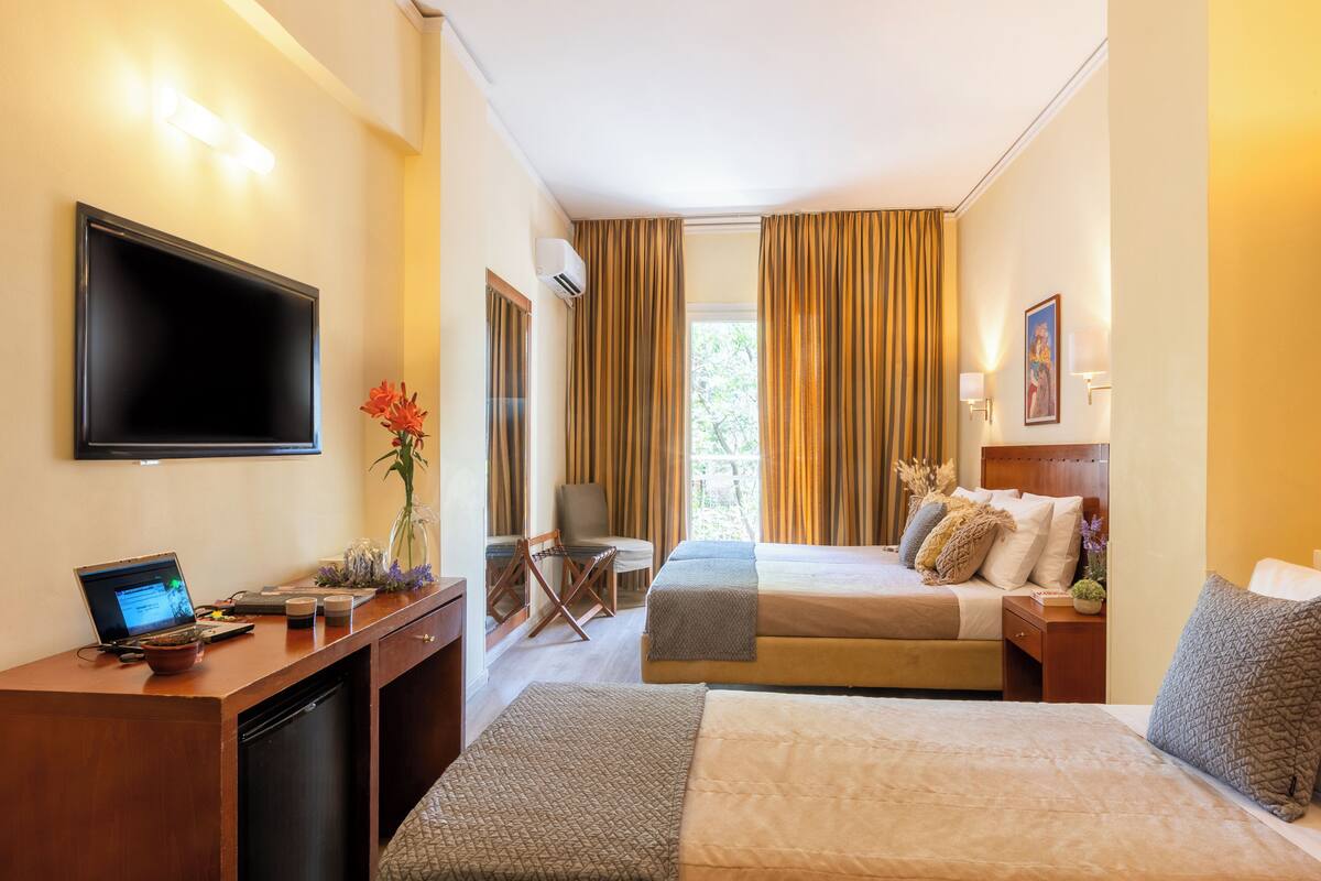 Quarto de hotel com duas camas, uma mesa de trabalho com um laptop, uma televisão e uma janela com cortinas.