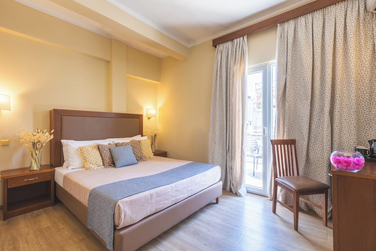 Quarto de hotel com uma cama grande, abajures, uma cadeira, uma escrivaninha e uma janela com cortinas.