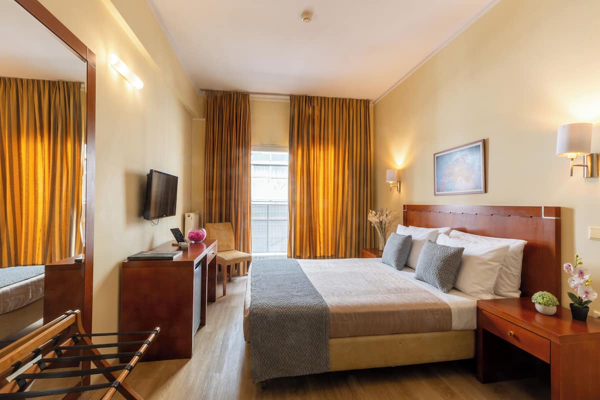Quarto de hotel com cama, mesa de cabeceira, televisão e escrivaninha com cadeira.