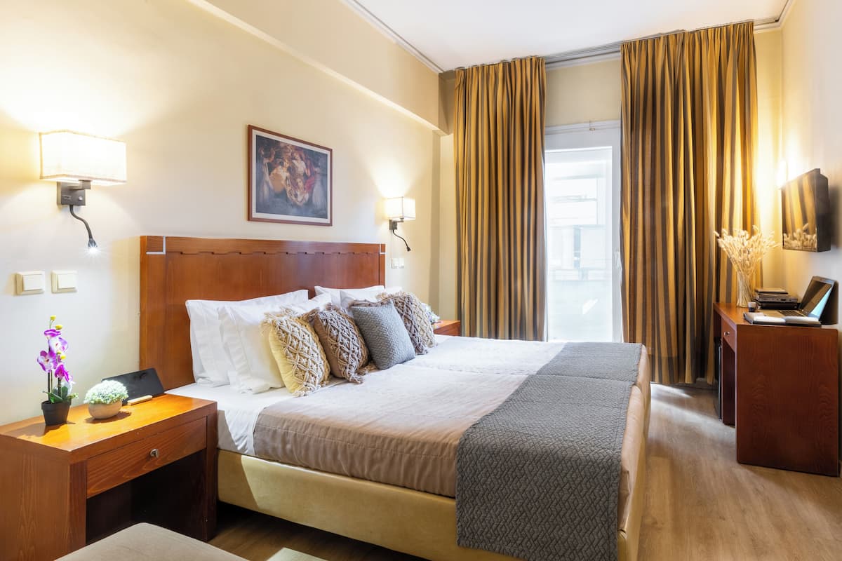 Quarto de hotel com uma cama grande, cabeceira de madeira, duas mesas de cabeceira com abajures, uma escrivaninha com um laptop e uma janela com cortinas.