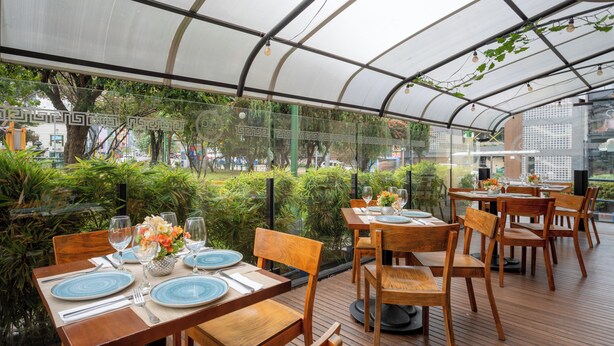 Um espaço de refeições interno com mesas e cadeiras, cercado por vegetação e um painel de vidro.