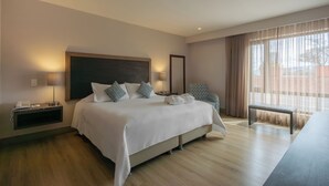 Quarto de hotel com uma cama grande, luminárias de cabeceira, telefone ao lado da cama, uma cadeira, um banco e uma janela com cortinas.