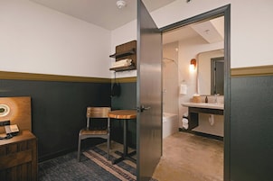 Quarto de hotel com uma cadeira de madeira, uma mesa pequena, um telefone, um porta-toalhas e um banheiro com pia e espelho.