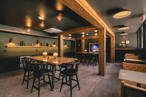 Interior de restaurante com mesa de jantar de madeira, cadeiras pretas e área de bar com banquetas.