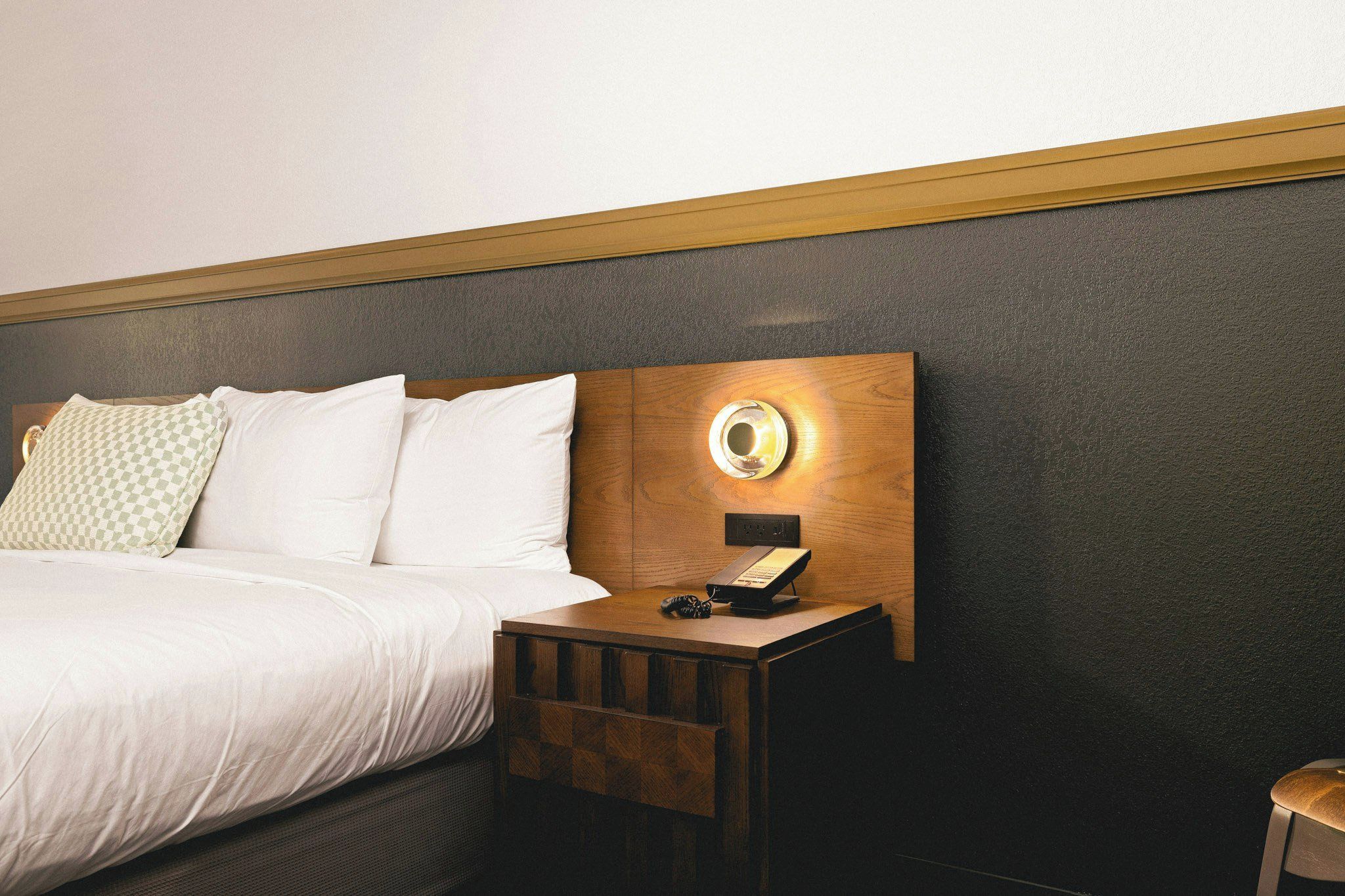 Quarto de hotel com cama, mesa de cabeceira com abajur e telefone fixado na parede.