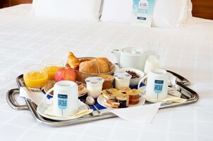 Bandeja de café da manhã com croissants, frutas, iogurte e café sobre a cama de um hotel.