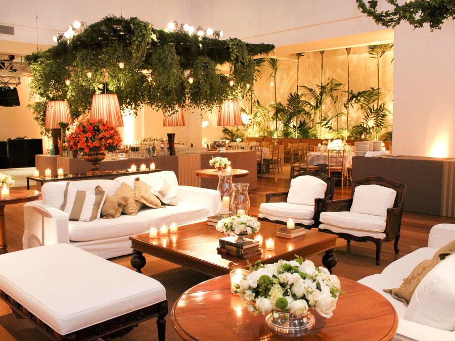 Uma área de estar bem iluminada e elegantemente decorada, com sofás e poltronas brancos e uma mesa de centro adornada com flores e velas.