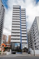 Um edifício moderno de vários andares com uma faixa vertical azul destacada, rodeado por outros edifícios.