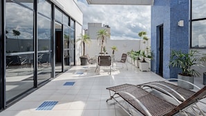 Um terraço no telhado com paredes de vidro, mesas ao ar livre e plantas em vasos.