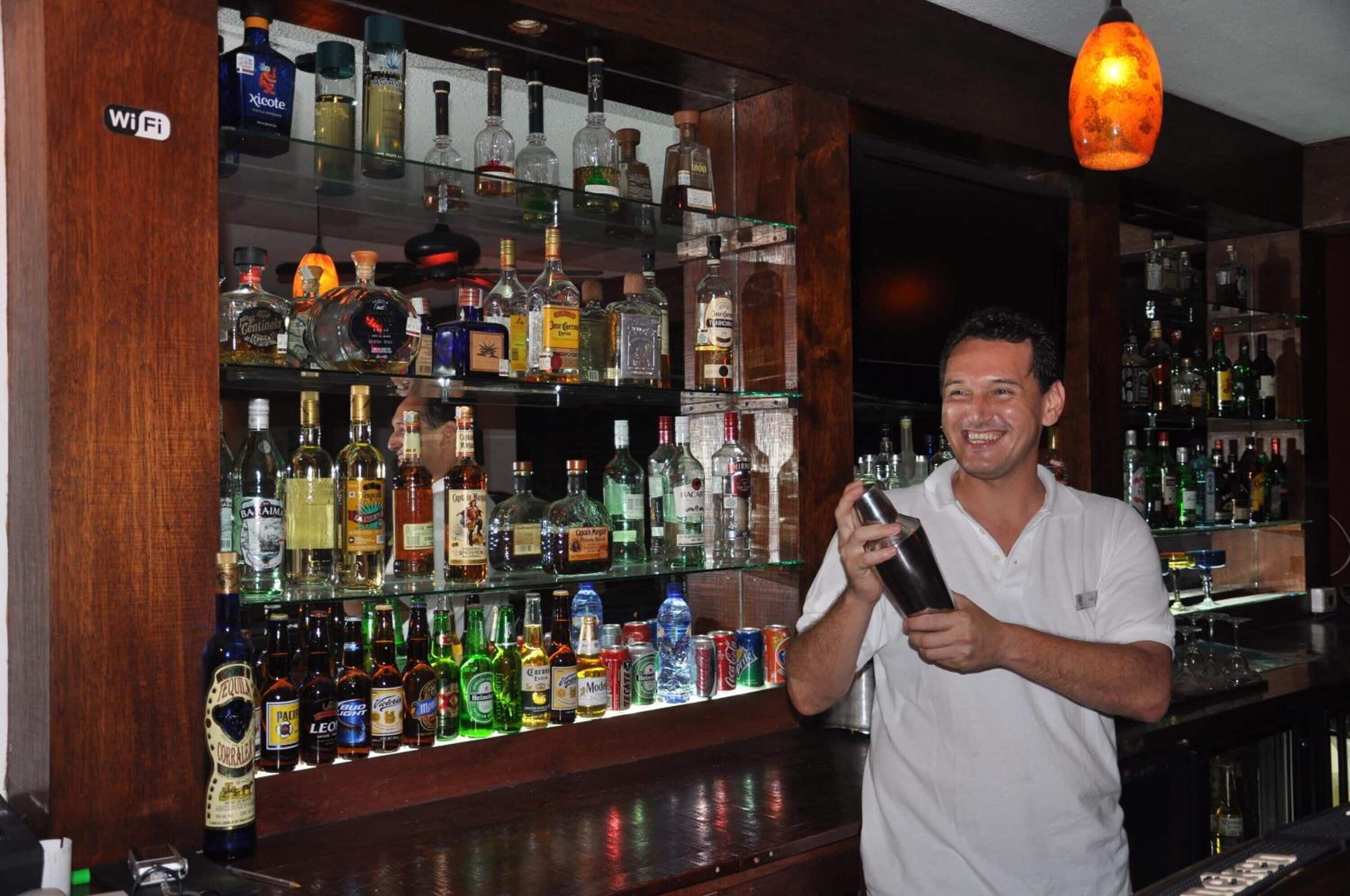Área de bar com prateleiras cheias de garrafas de bebidas e uma pessoa segurando um coqueteleira.
