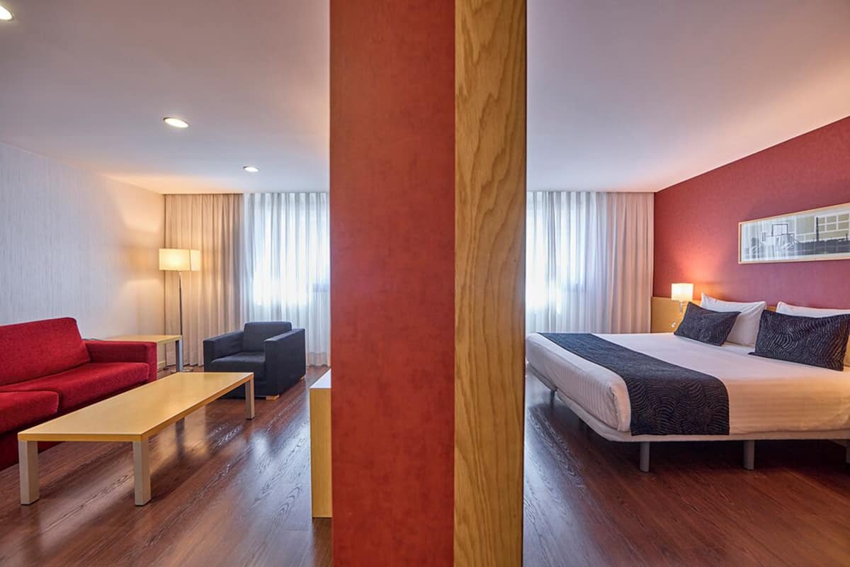 Quarto de hotel com cama, sofá vermelho, mesinha e uma cadeira.