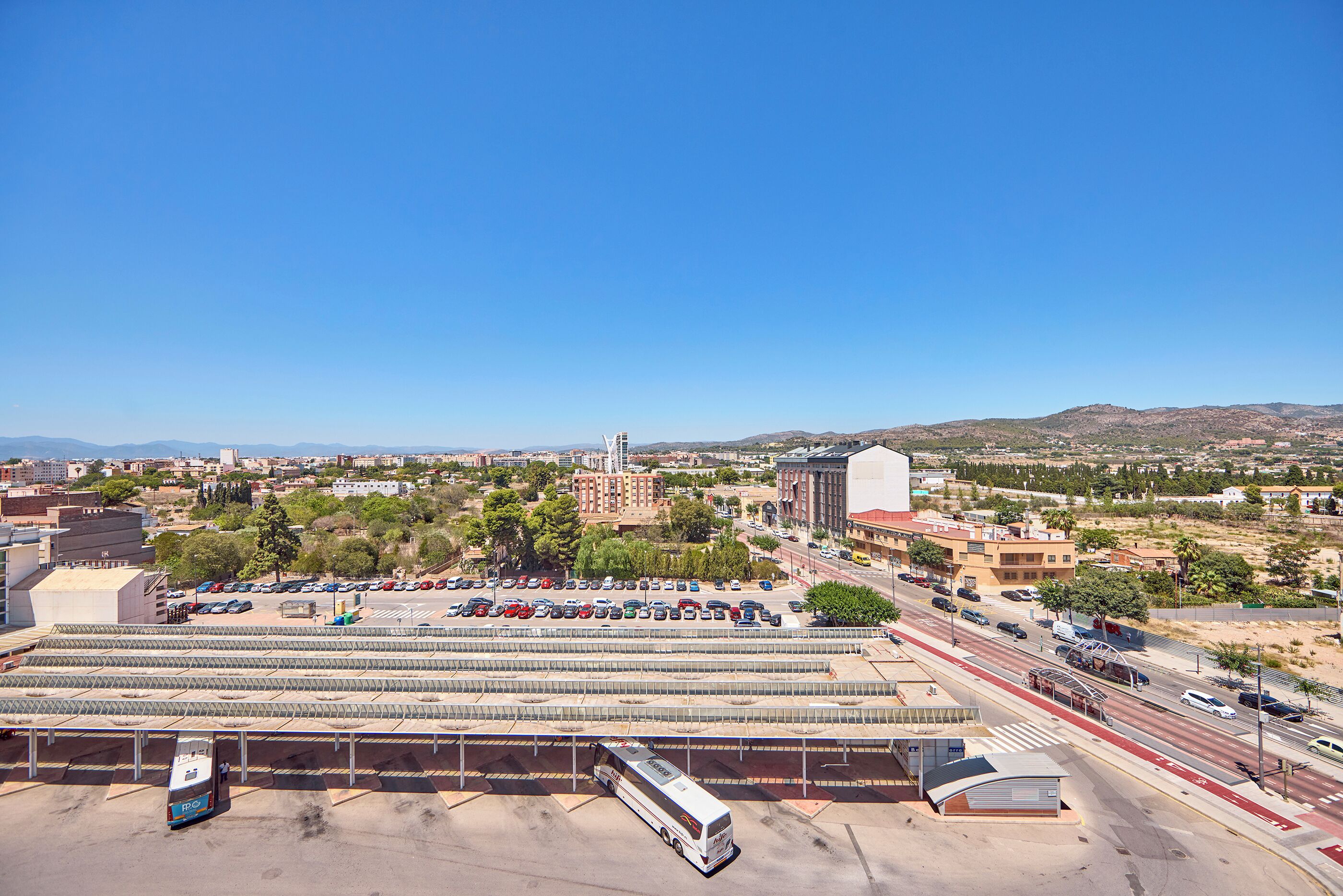 Uma rodoviária com vários ônibus, um estacionamento e vista para um horizonte urbano com edifícios e colinas.