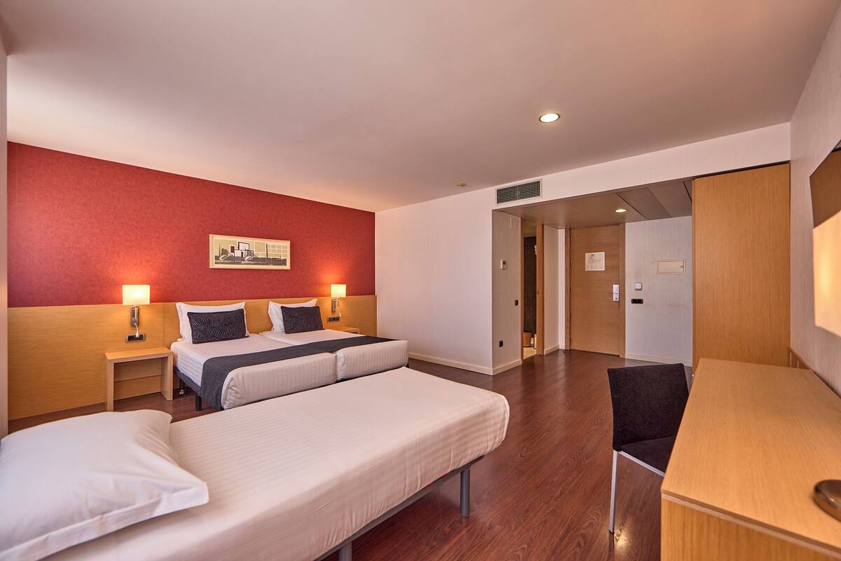 Quarto de hotel com duas camas, uma escrivaninha e uma cadeira. Uma parede vermelha com um quadro e uma porta de madeira ao fundo.