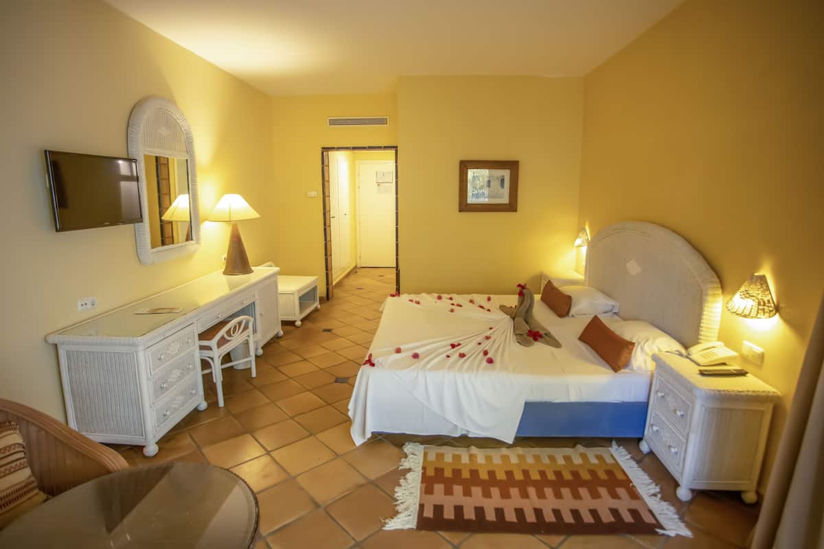 Quarto de hotel com cama, mesas de cabeceira, escrivaninha, cadeira, televisão e porta para outro cômodo.