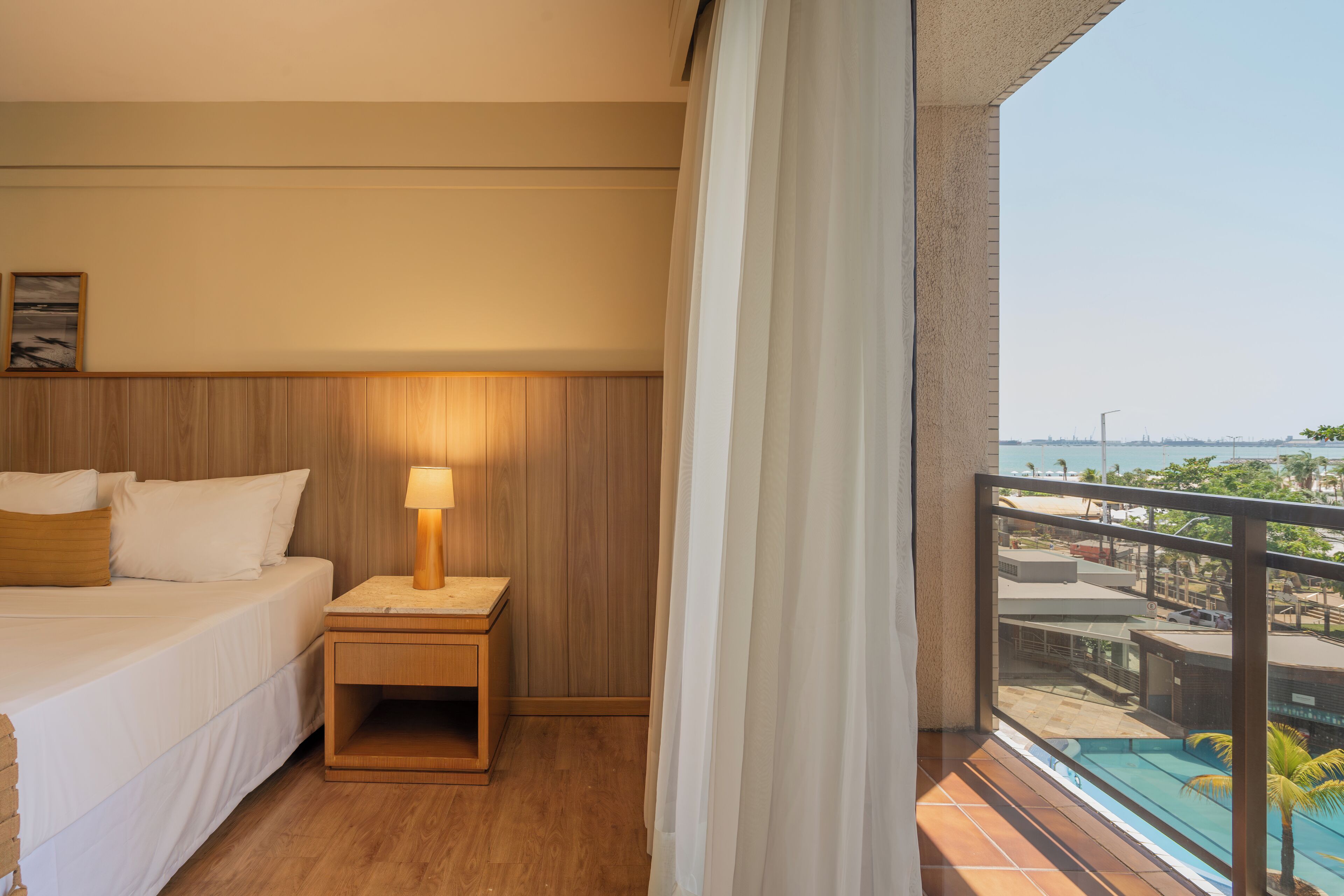 Quarto de hotel com cama, cabeceira de madeira, mesa de cabeceira com abajur e varanda com vista para a piscina.