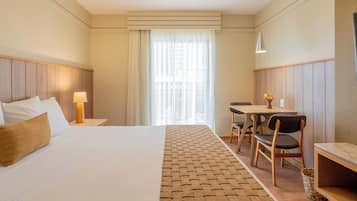 Quarto de hotel com uma cama grande, uma escrivaninha com cadeira, um abajur e uma janela com cortinas finas.