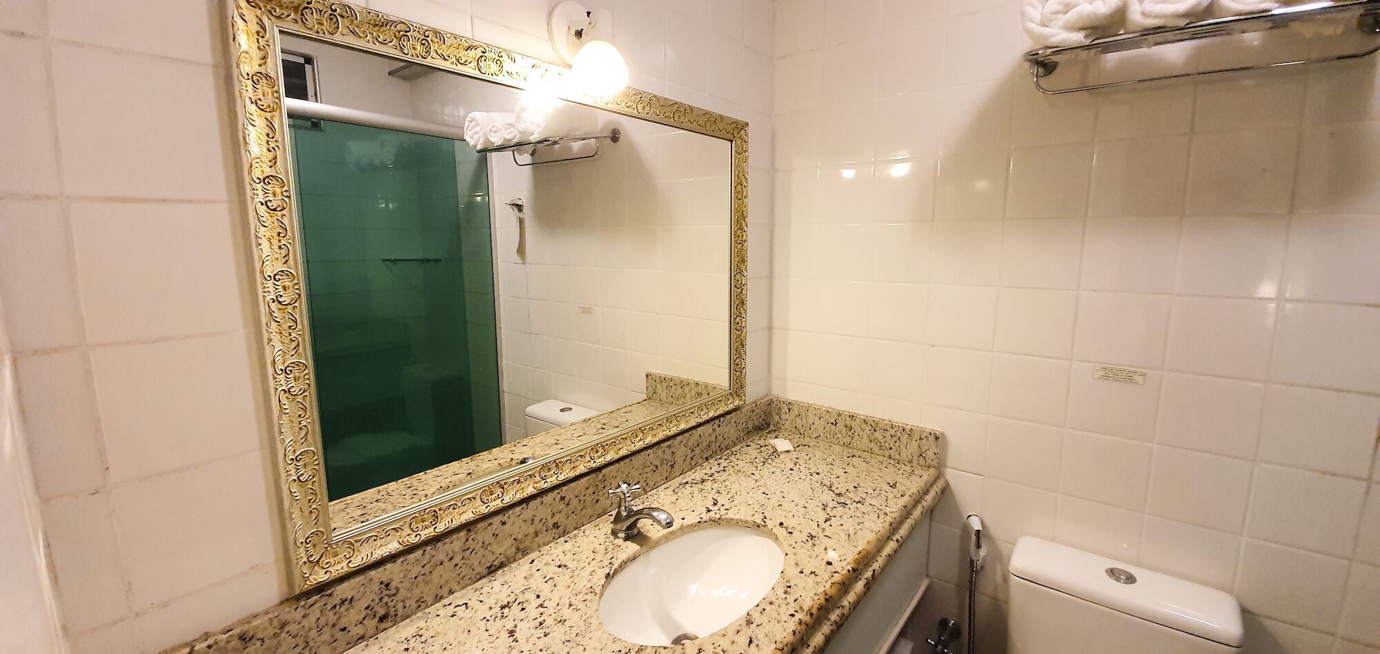 Casa de banho com lavatório bancada de granito, espelho com moldura dourada e sanita branca.