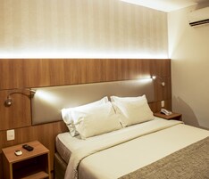 Quarto de hotel com cabeceira de madeira, cama com lençóis brancos, mesa de cabeceira com um controle remoto e um telefone.