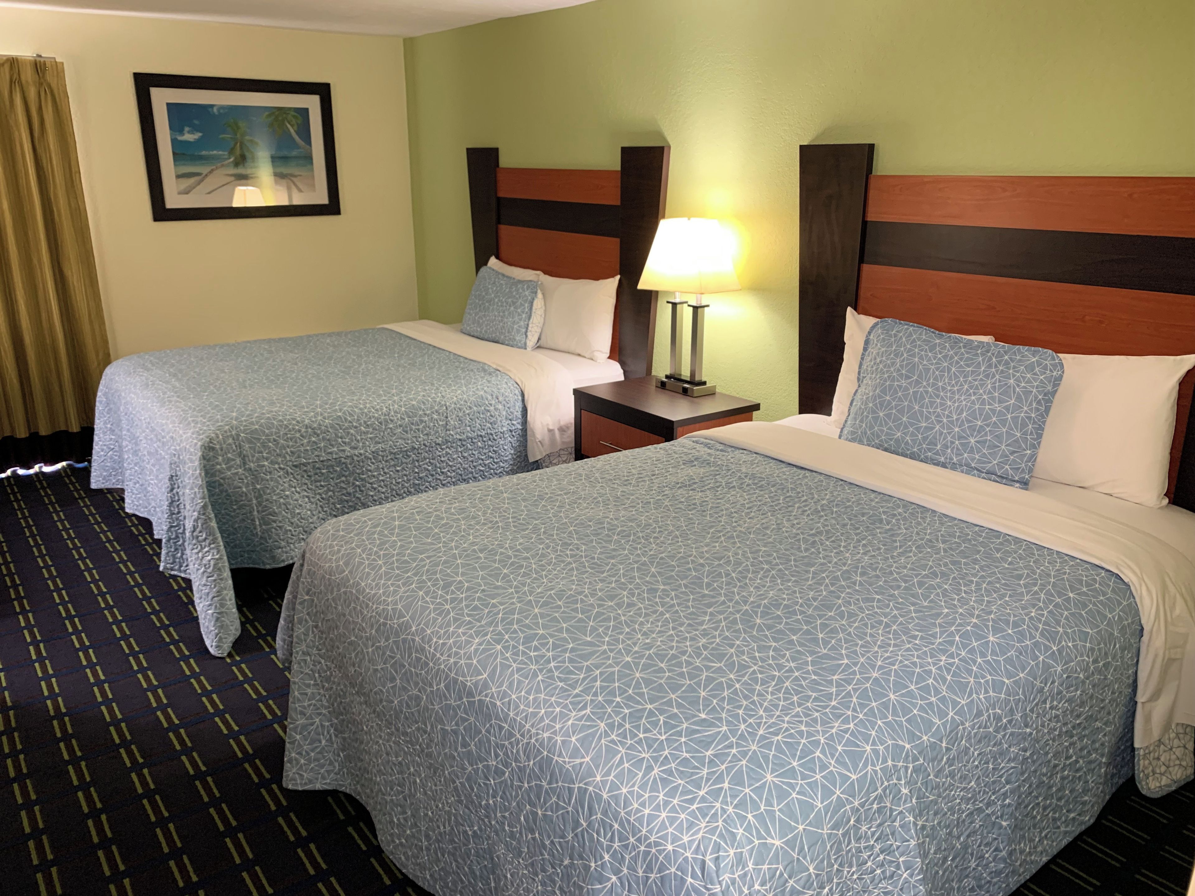 Quarto de hotel com duas camas, uma mesa de cabeceira com abajur, um quadro na parede e um tapete estampado.