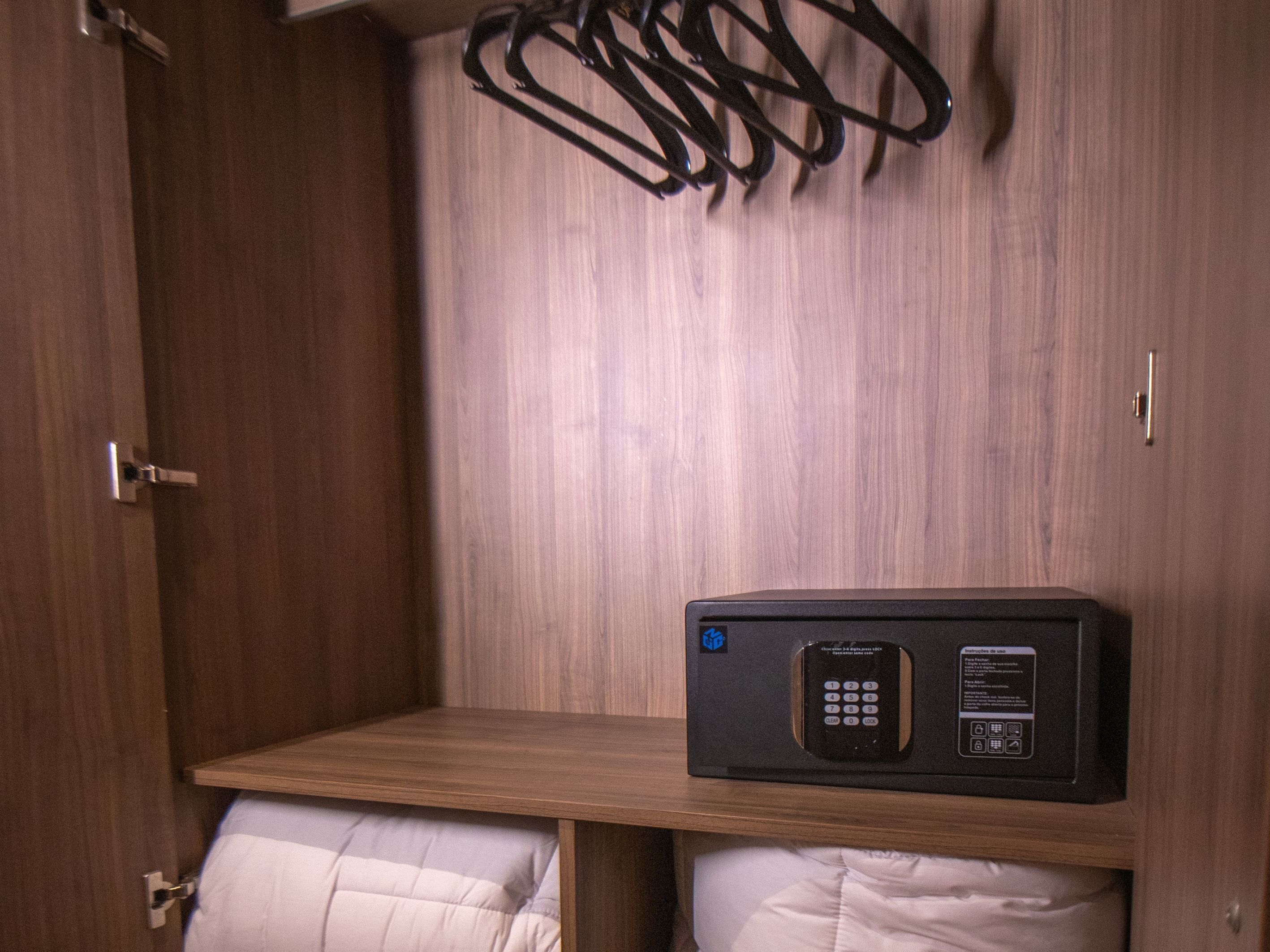 Quarto compacto com guarda-roupa de madeira, uma cama pequena com colchão branco e um dispositivo eletrônico preto sobre uma prateleira de madeira.