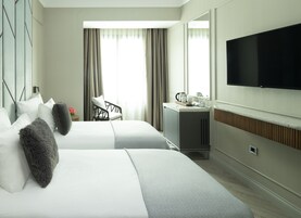 Quarto de hotel com duas camas, uma escrivaninha com chaleira, uma cadeira, televisão de tela plana e janela com cortinas.