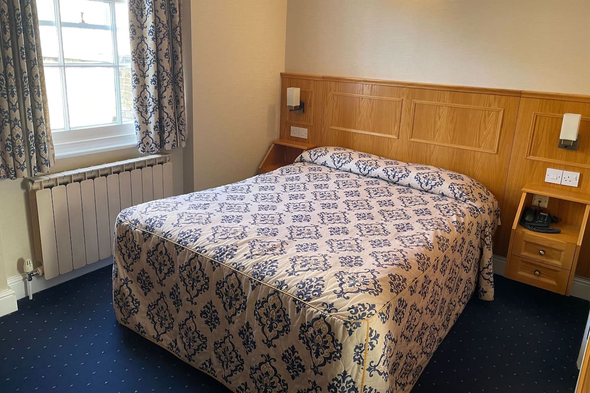 Quarto de hotel com uma cama, mesa de cabeceira com telefone, luminária na parede e uma janela com cortinas.
