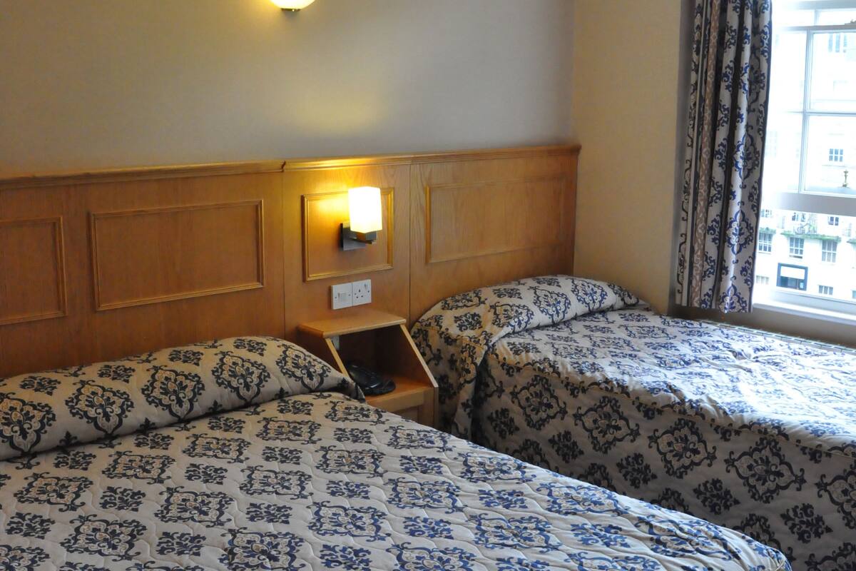 Quarto de hotel com duas camas, mesa de cabeceira, luminária na parede e janela com cortinas.