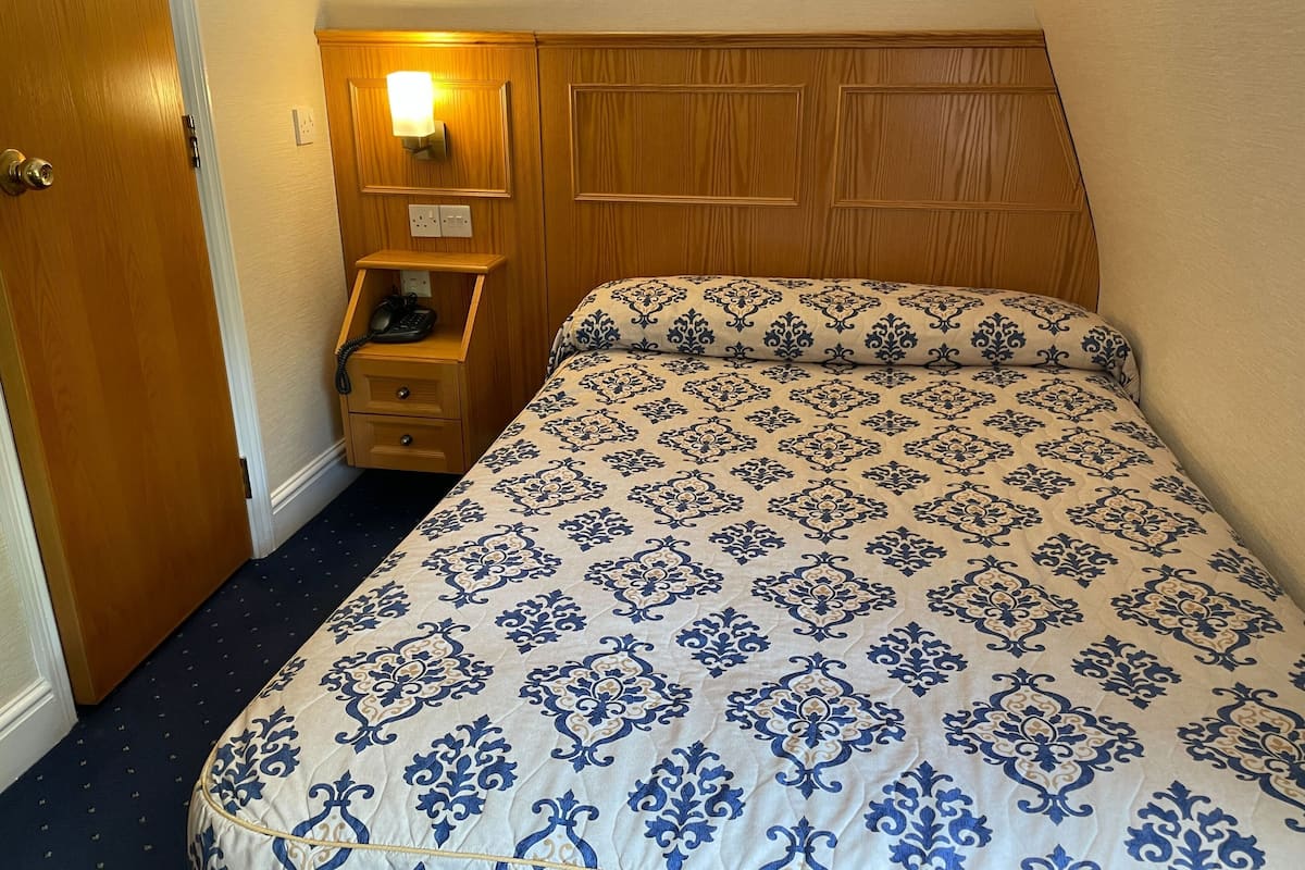 Quarto de hotel com cama, mesa de cabeceira de madeira, telefone e luminária fixa na parede.
