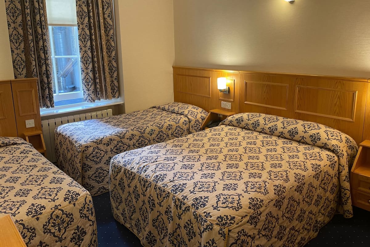 Quarto de hotel com duas camas de solteiro, janela com cortinas, luminária fixada na parede e radiador.