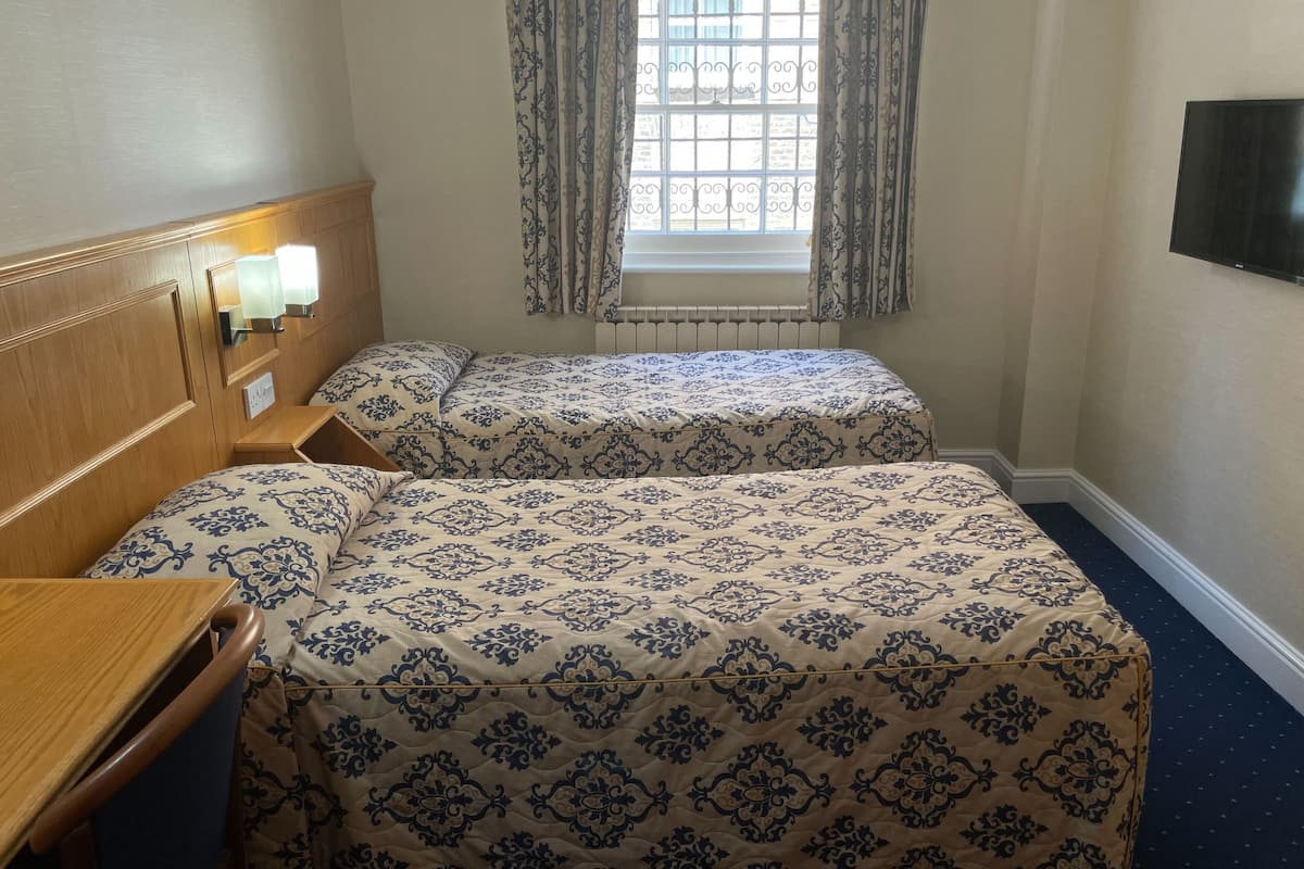 Quarto de hotel com duas camas, cabeceira de madeira, escrivaninha, janela com cortinas e televisão na parede.