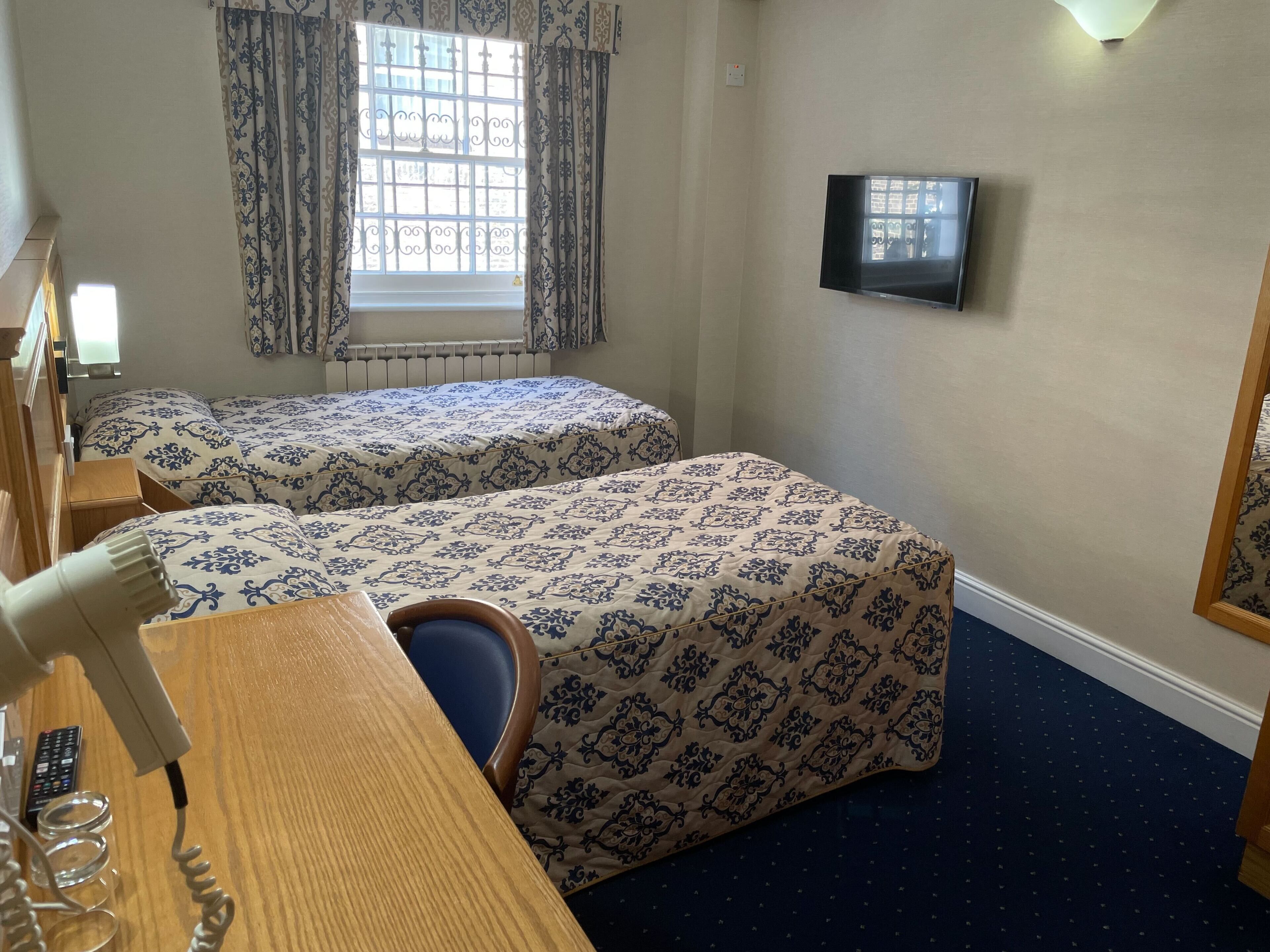 Quarto de hotel com duas camas, uma mesa com telefone, uma cadeira, um radiador, televisão na parede e uma janela com cortinas.